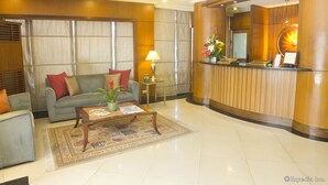 Um hall de hotel com uma mesa de recepção, um sofá, uma mesa de centro e uma pequena mesa lateral com uma luminária.