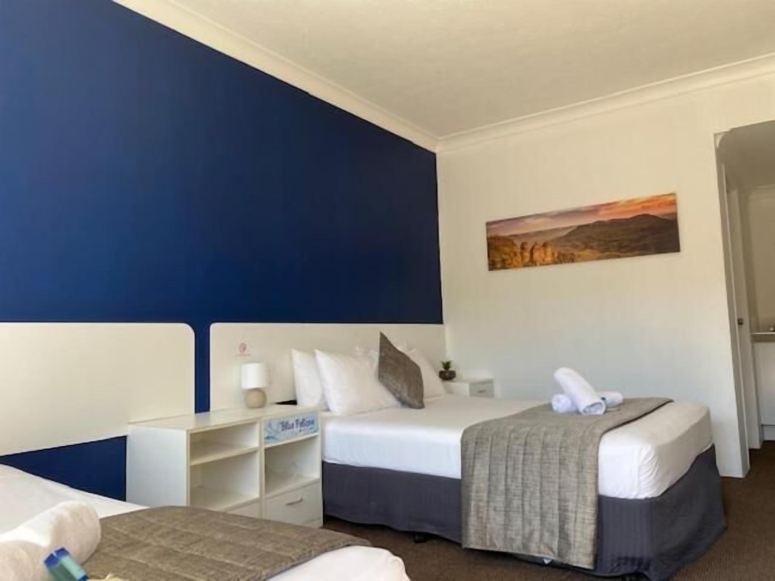 Quarto de hotel com duas camas, uma mesa de cabeceira, uma mesinha e um quadro na parede.