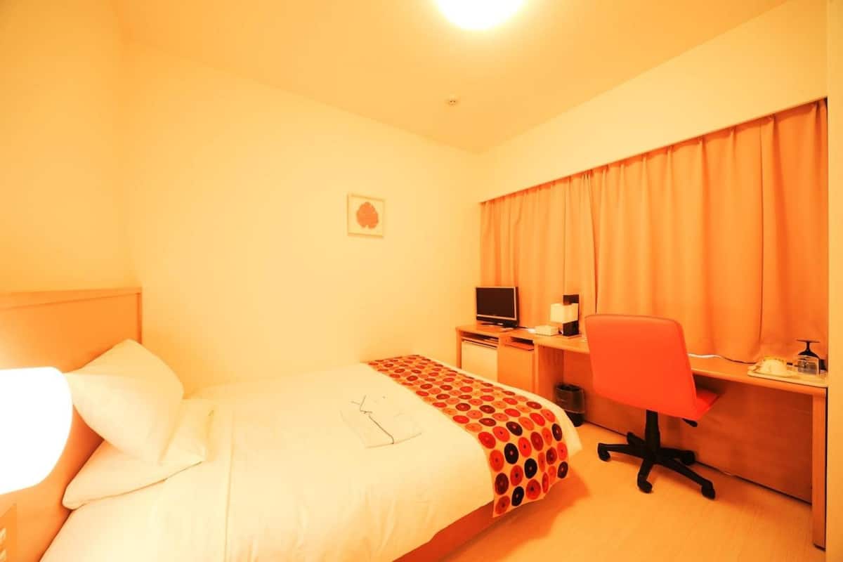 Quarto de hotel com cama, escrivaninha, cadeira, televisão e janela com cortinas.