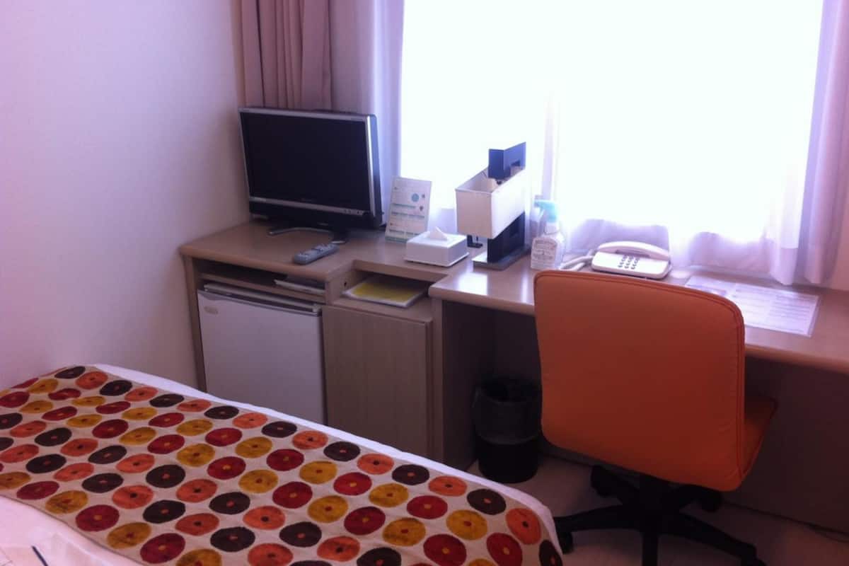 Quarto de hotel com cama, escrivaninha com computador, telefone e janela com cortinas.