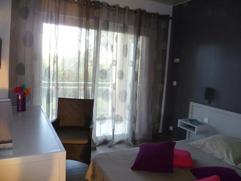 Quarto de hotel com cama, uma cadeira, escrivaninha e janela com cortinas.