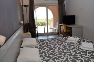 Quarto de hotel com cama, escrivaninha com televisão, cadeira e vista para uma varanda com mobiliário exterior.