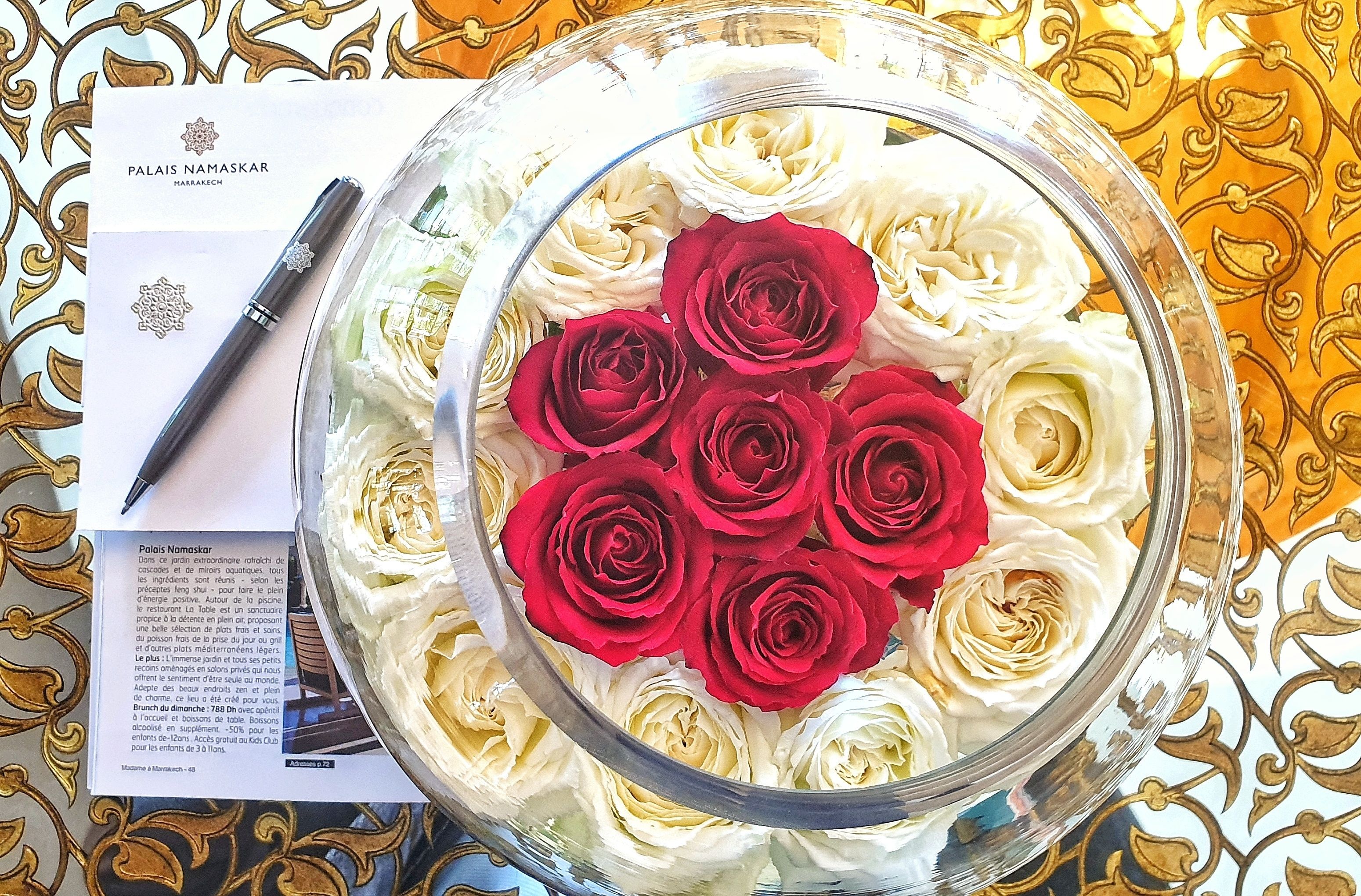 Uma tigela de vidro com rosas vermelhas e brancas, uma caneta e um folheto sobre uma toalha de mesa estampada.