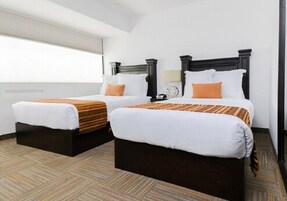 Quarto de hotel com duas camas, cada uma com cabeceira escura, roupa de cama branca e almofadas laranja. O quarto tem piso de carpete e uma janela grande.