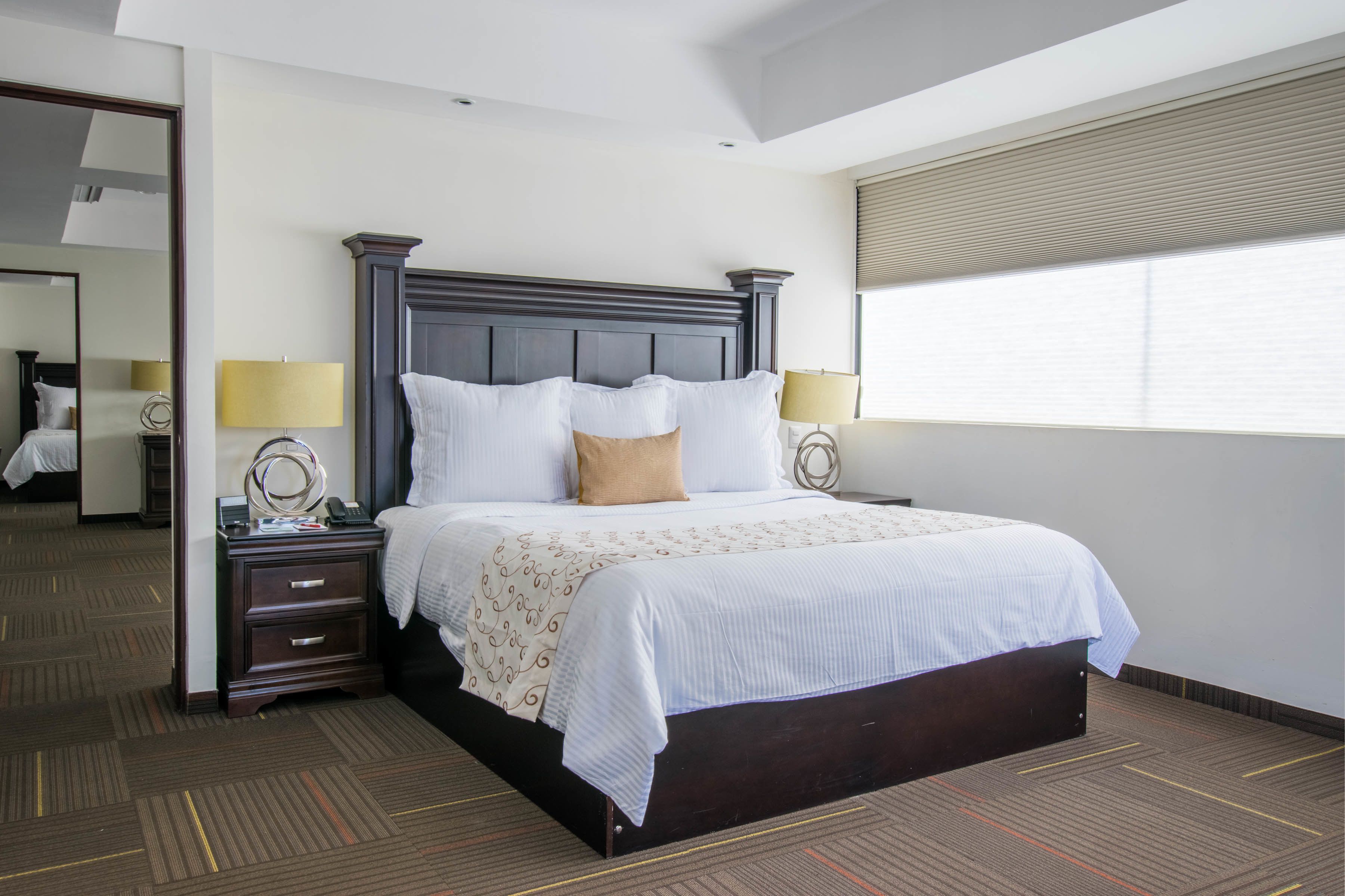 Quarto de hotel com uma cama grande, duas mesas de cabeceira, um espelho e uma janela com persianas.