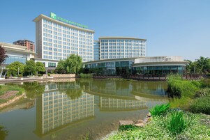 Hotel moderno refletido em um lago, cercado por vegetação e céu claro.