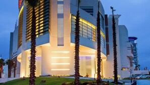 Um hotel moderno com um letreiro chamativo da "Fiesta Inn", vários andares e uma entrada bem iluminada.