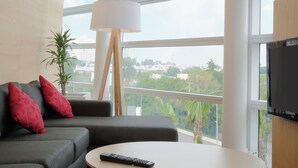 Uma sala de estar moderna com um sofá cinzento, almofadas vermelhas, uma mesa redonda, um comando de televisão, um candeeiro de pé e uma TV sobre um suporte de madeira.