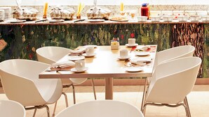 Uma área de refeições com uma mesa comprida posta para várias pessoas, rodeada por cadeiras brancas.