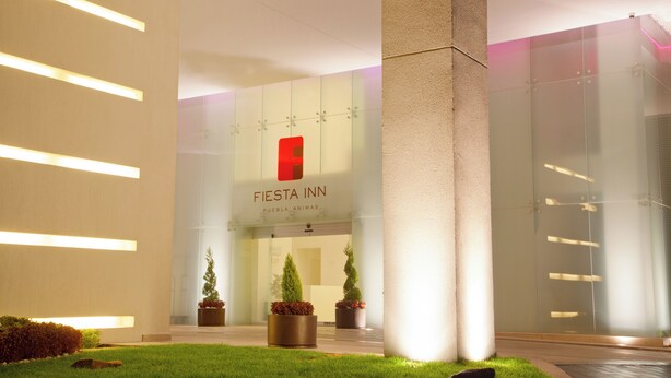 Exterior do Fiesta Inn, com design moderno, apresentando fachada de vidro, letreiros iluminados e vasos de plantas em primeiro plano.