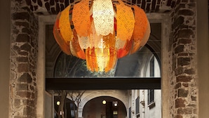 Um corredor amplo com um lustre grande e ornamentado, paredes de tijolo e uma área de estar com mesas e cadeiras.