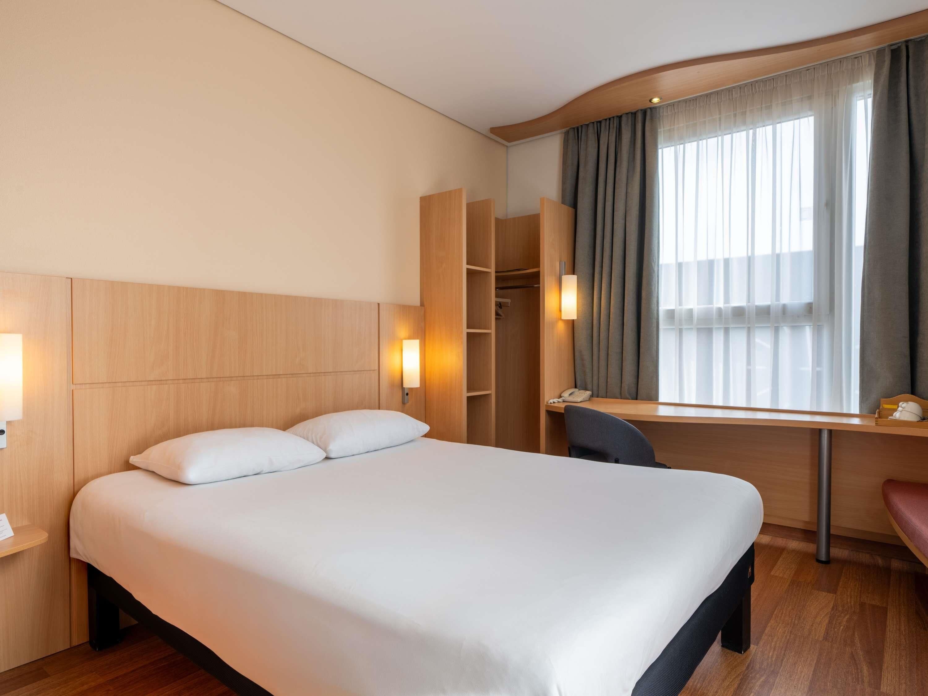 Quarto de hotel com uma cama grande, uma escrivaninha com cadeira, um guarda-roupa e uma janela com cortinas.