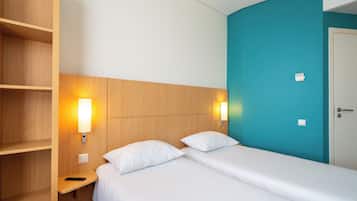 Quarto de hotel com uma cama, duas mesas de cabeceira, um criado-mudo, uma luminária fixada na parede e uma porta.