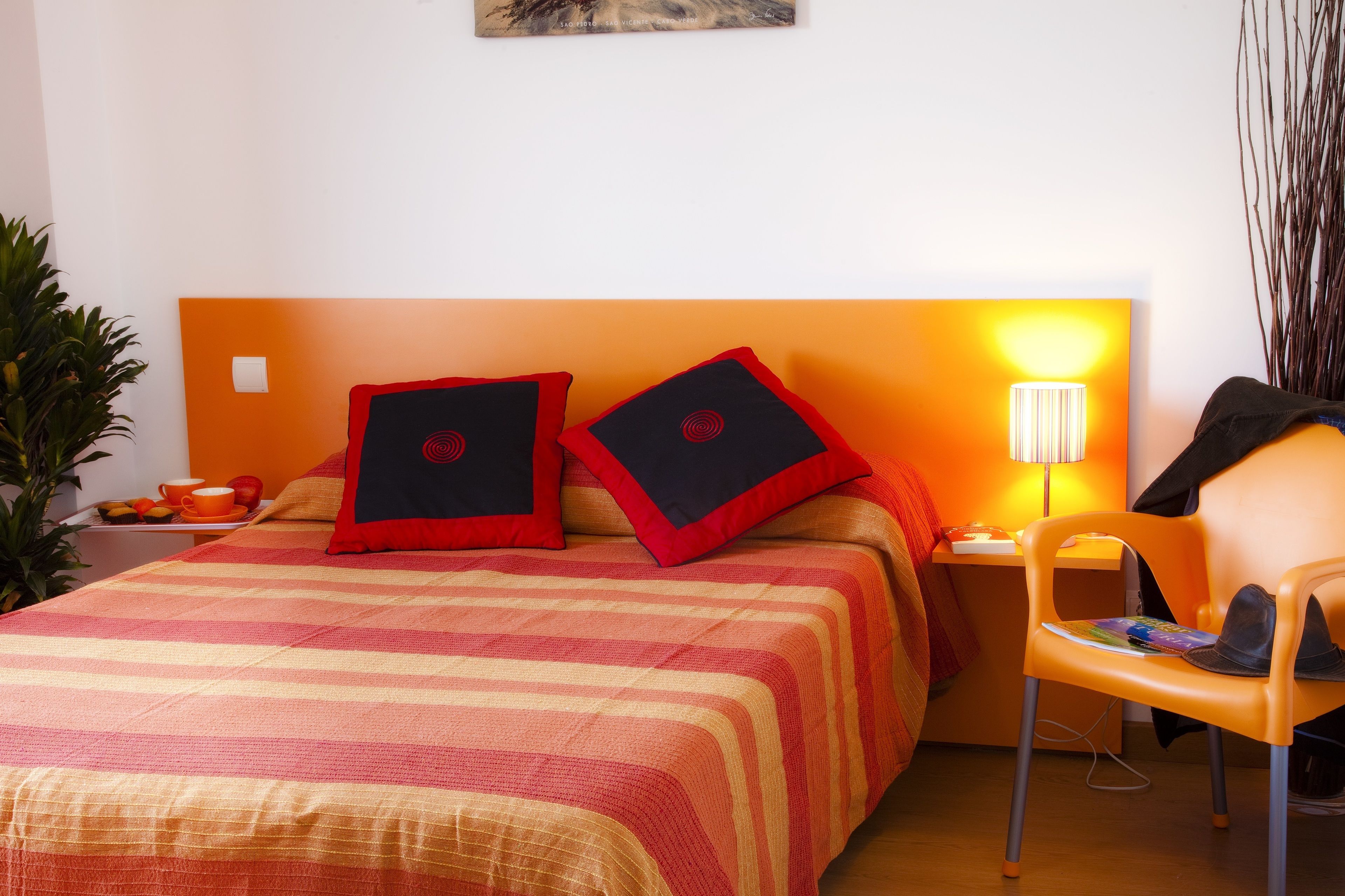 Quarto de hotel com cama, duas almofadas, mesa de cabeceira, luminária, cadeira e planta.