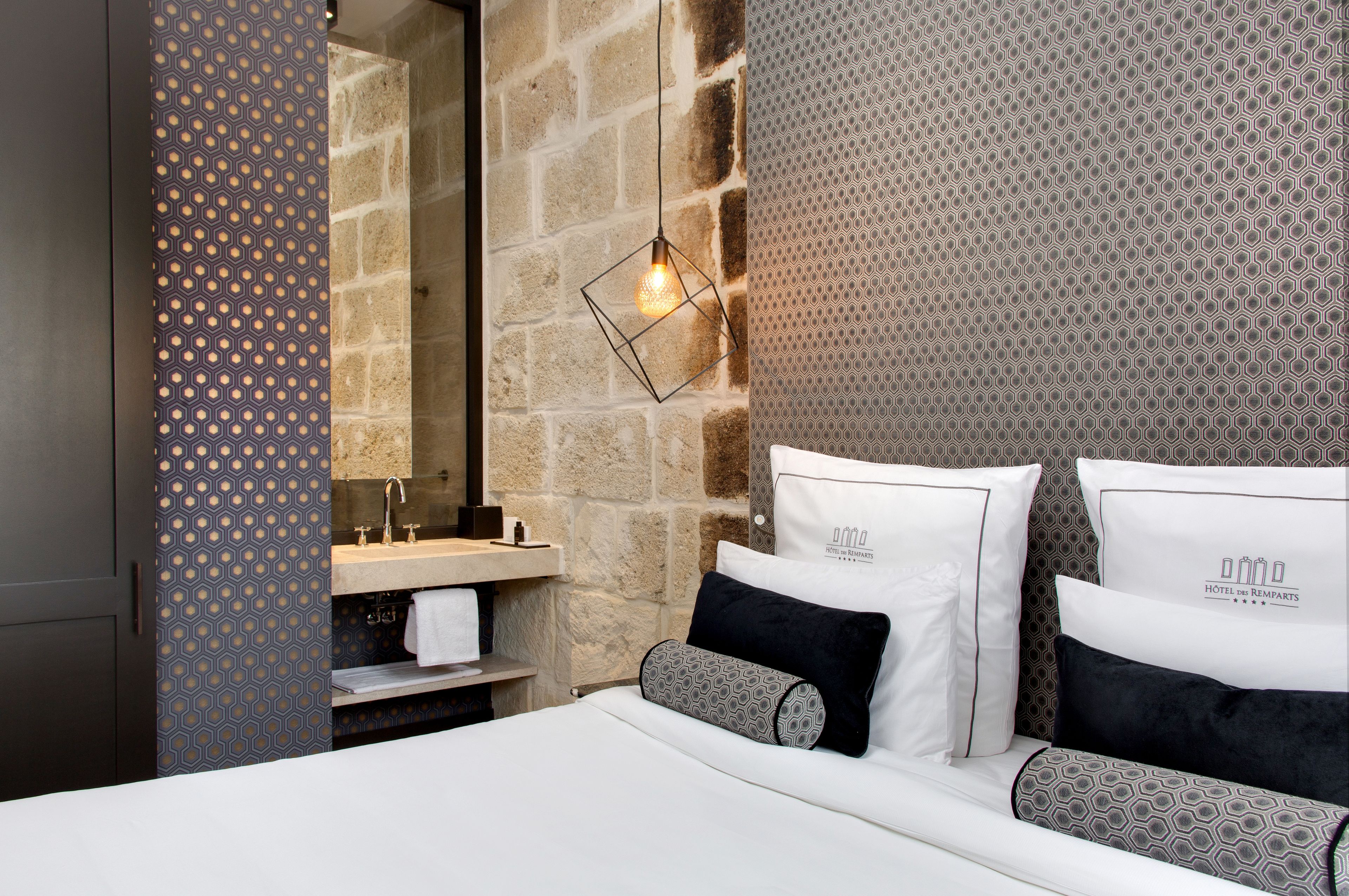 Quarto de hotel com parede de pedra, luminária moderna, cama com almofadas estampadas e banheiro com espelho e pia.