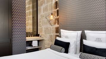 Quarto de hotel com parede de pedra, luminária moderna, cama com almofadas estampadas e banheiro com espelho e pia.