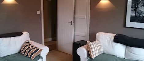 Um quarto com duas sofás brancas, cada uma com almofadas de estampas diferentes, uma manta verde e uma mesinha de cabeceira preta. Há luminárias fixadas na parede, de cada lado de uma porta, e um quadro pendurado na parede.
