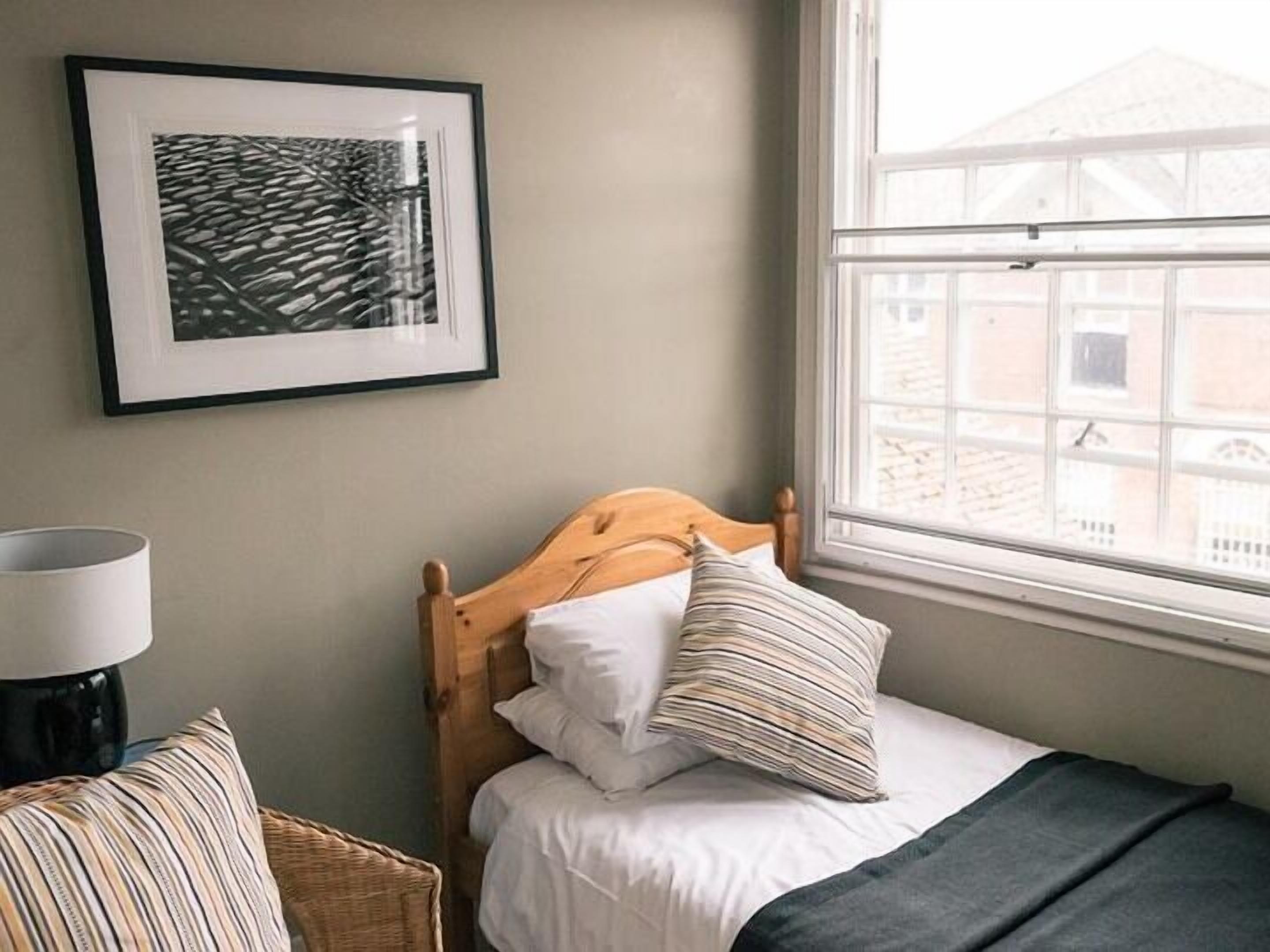 Quarto com cama de madeira, roupa de cama branca, almofada listrada, abajur preto e quadro na parede.