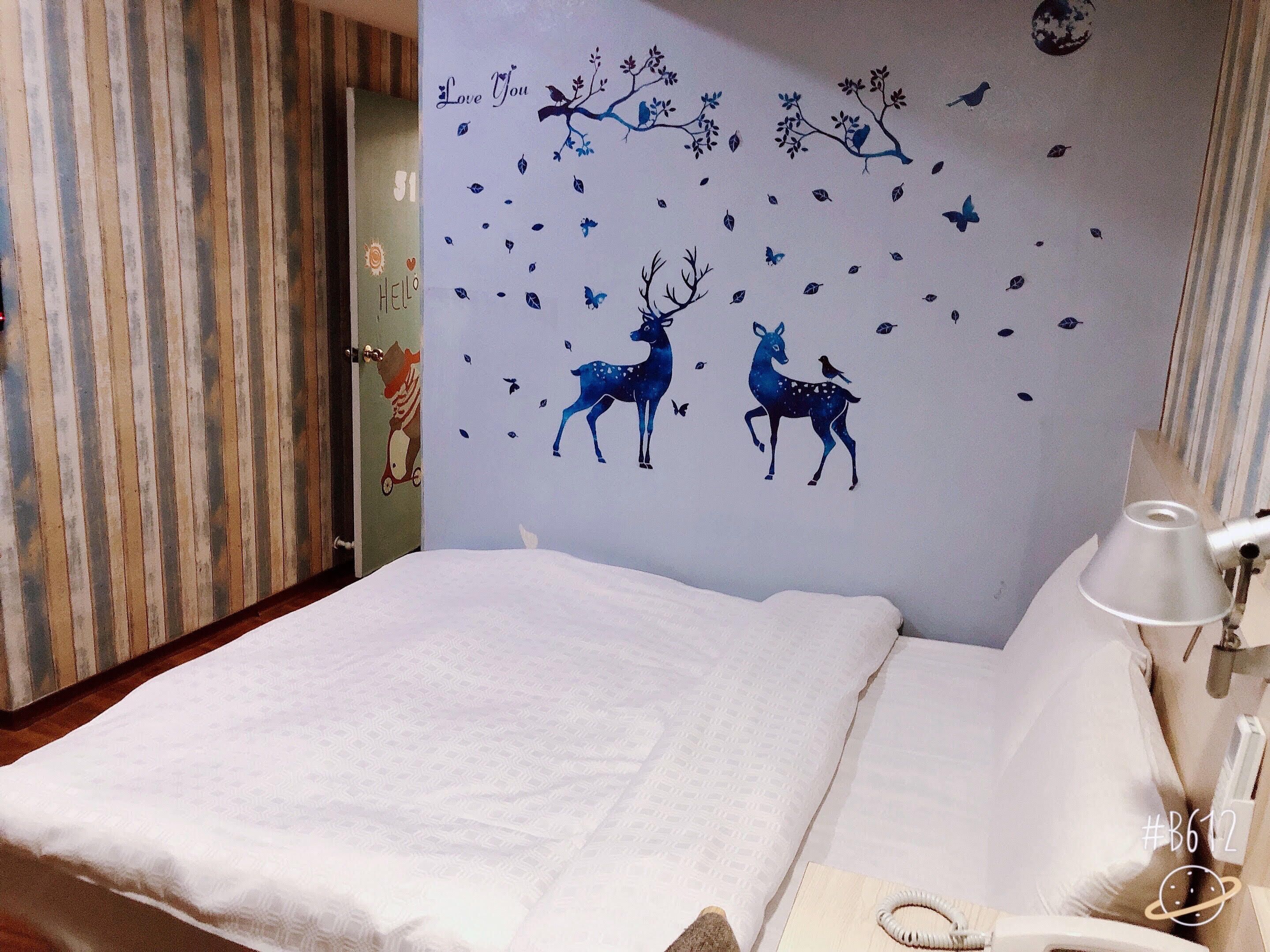 Quarto de hotel com cama, mesa de cabeceira, abajur e parede decorada com um decalque de tema natural.