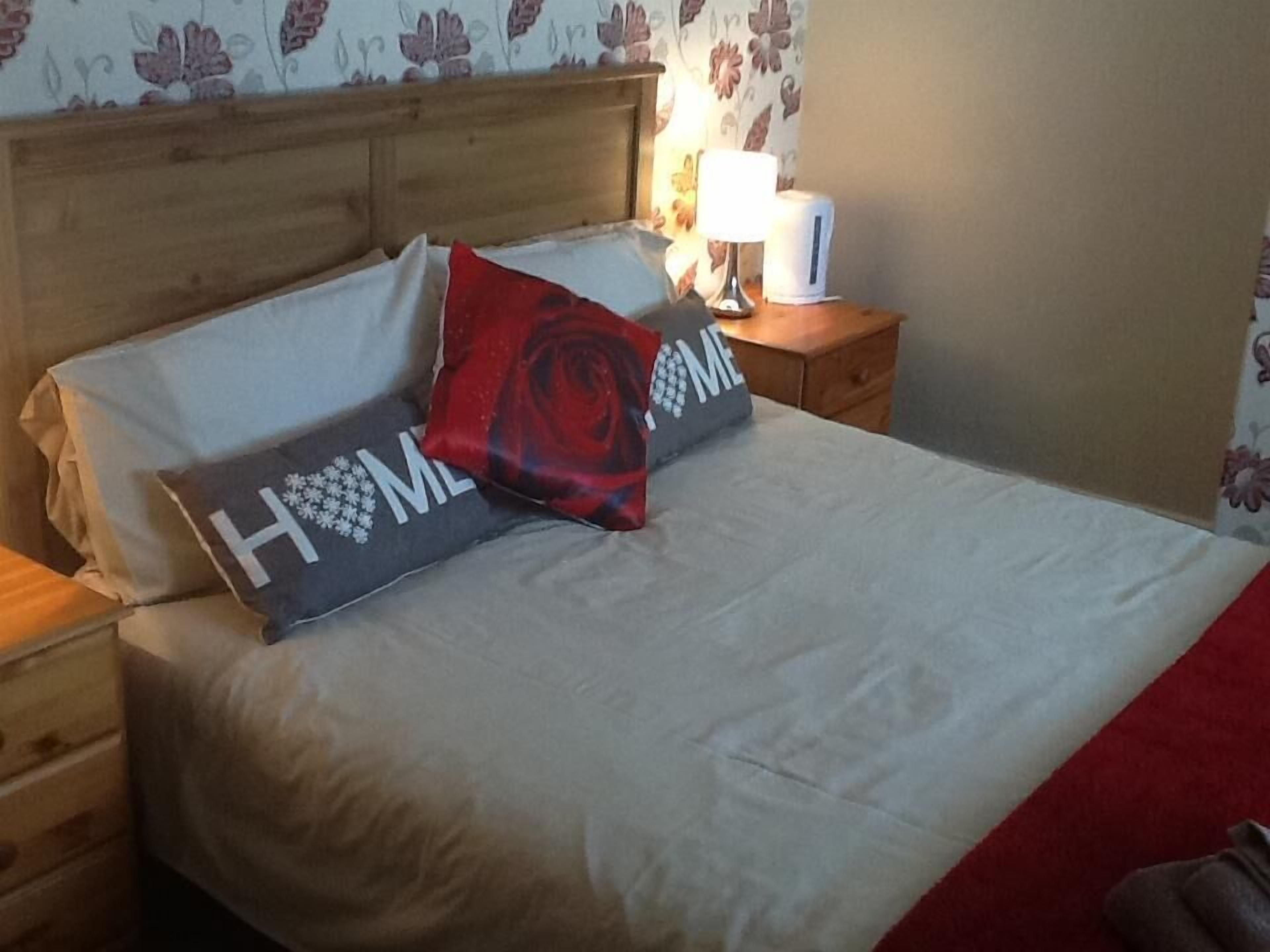 Cama com duas almofadas, uma estampada com padrão floral e outra com desenho de rosa vermelha. Há uma mesa de cabeceira com abajur e um pequeno aparelho branco.
