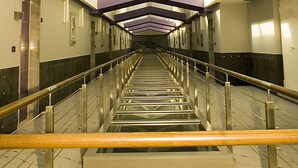 Um corredor comprido e estreito com um eixo de elevador de vidro, grades de metal e uma série de portas em ambos os lados.