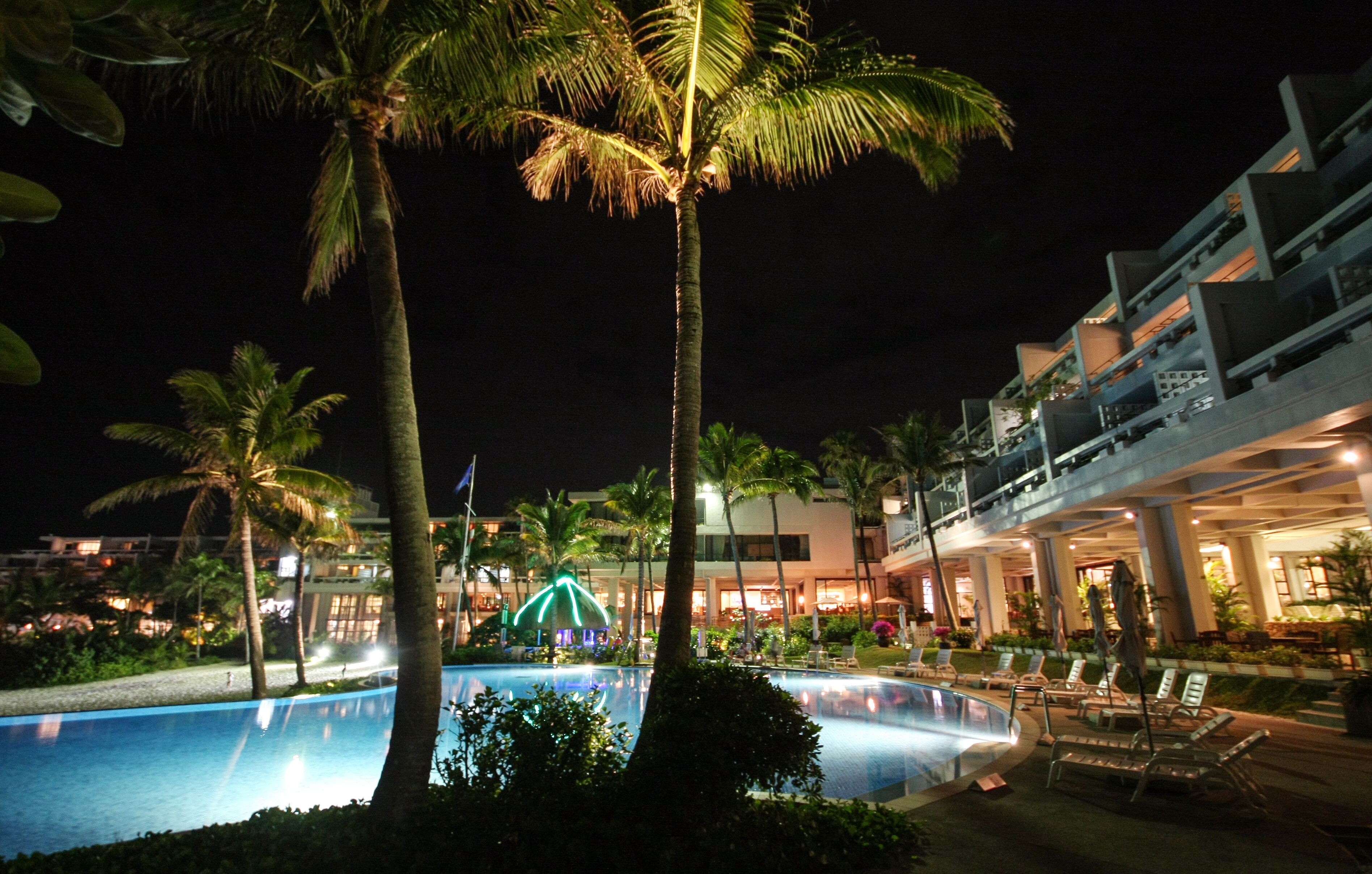 Vista noturna de uma área de piscina de hotel, com palmeiras, espreguiçadeiras e um edifício com varandas.