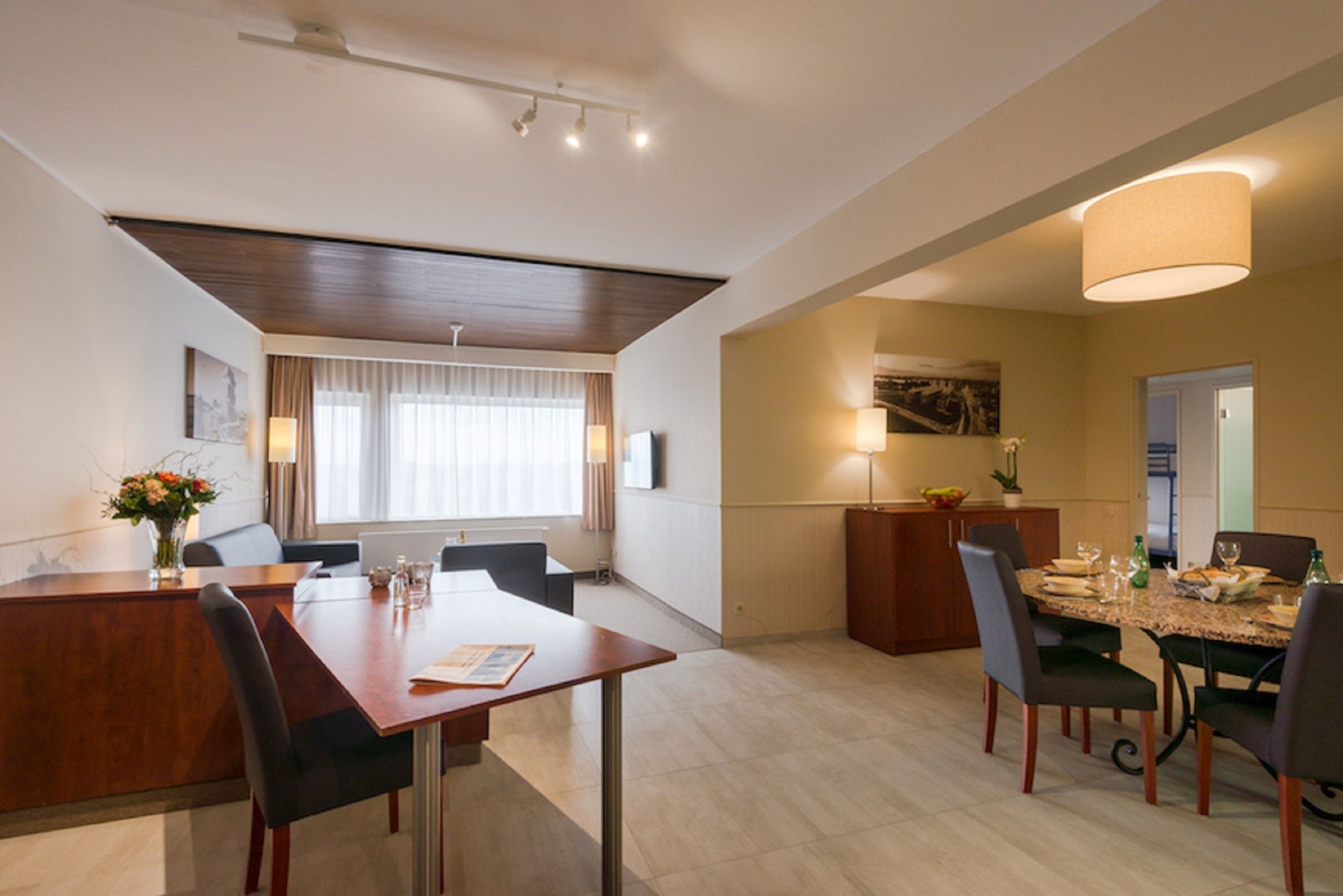 Quarto de hotel moderno com área de refeições, escrivaninha de madeira e uma janela grande com cortinas.