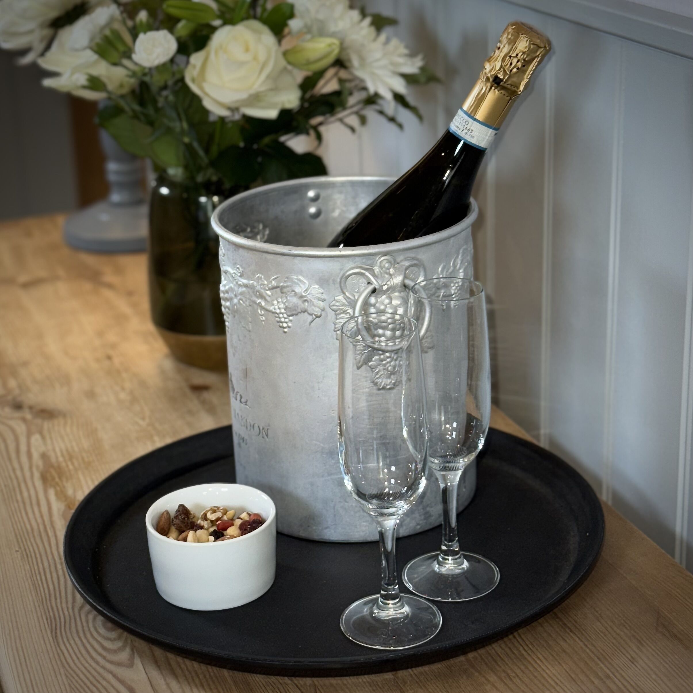 Uma garrafa de champanhe em um balde de metal decorativo, com duas taças, uma pequena tigela de castanhas e um vaso de flores sobre uma mesa de madeira.