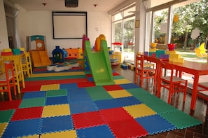 Uma sala de brinquedos com tapetes de espuma coloridos, um escorrega e diversas estruturas lúdicas.