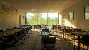 Uma sala de conferências com várias mesas e cadeiras, amplas janelas com vista para vegetação e teto com padrão quadriculado.