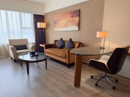 Quarto de hotel moderno com sofá, duas poltronas, mesa de centro de vidro e luminária.