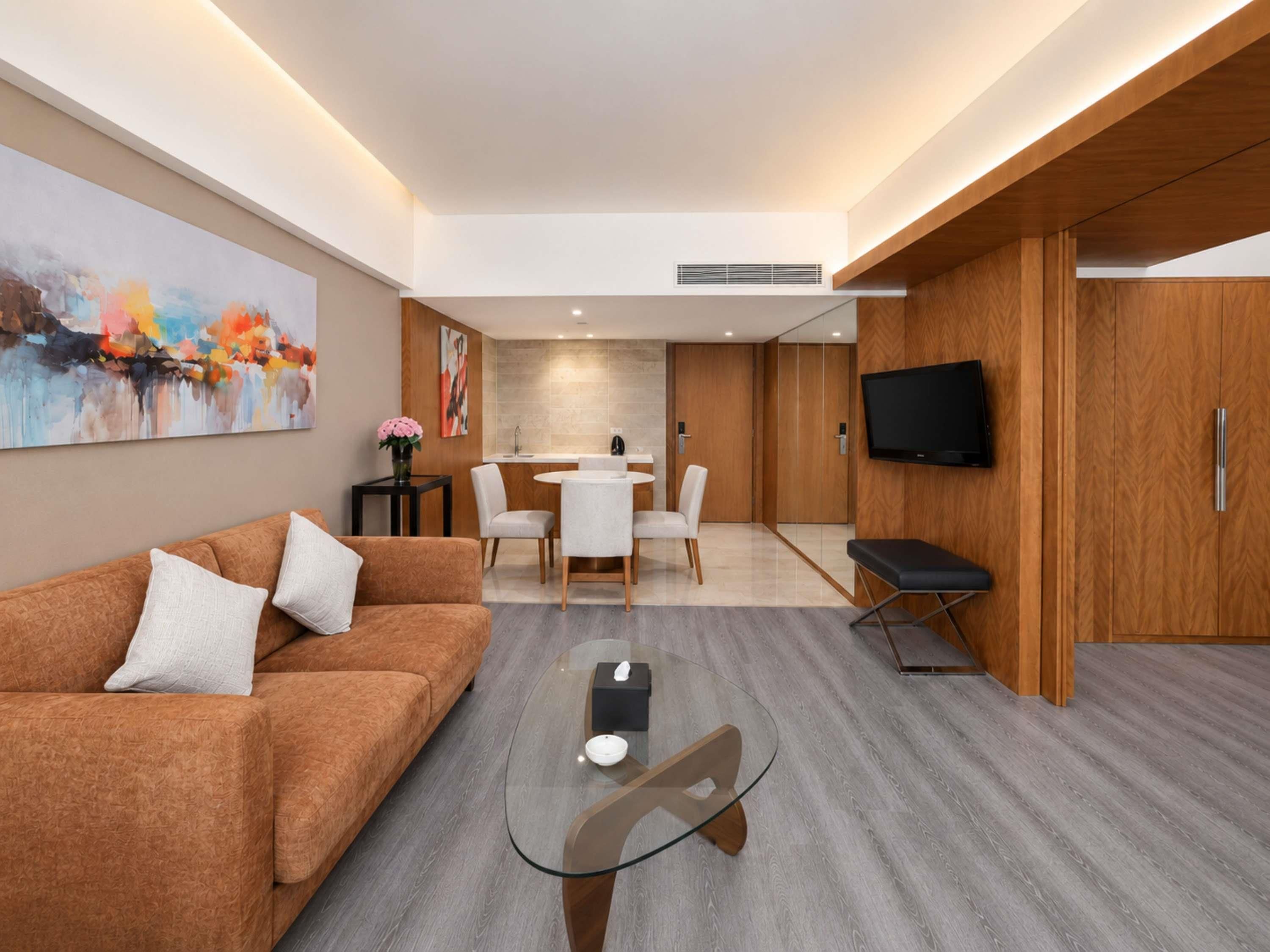 Uma sala de estar moderna com sofá, mesinha de centro de vidro, área de jantar com cadeiras, cozinha e televisão fixada numa parede de madeira.