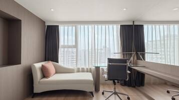 Quarto de hotel moderno com sofá curvo, escrivaninha com cadeira e janelas amplas com vista para a cidade.
