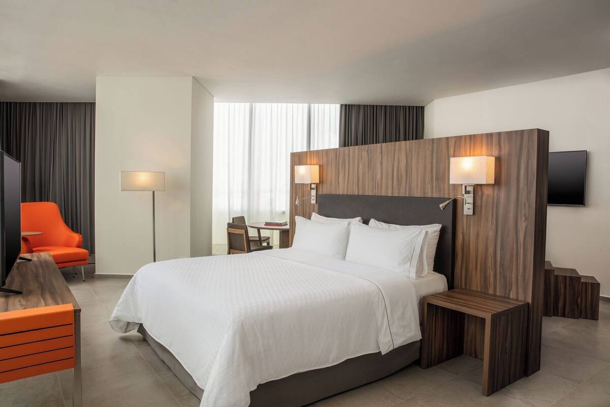 Quarto de hotel com uma cama grande, uma escrivaninha, uma cadeira laranja, uma televisão e uma janela com cortinas.