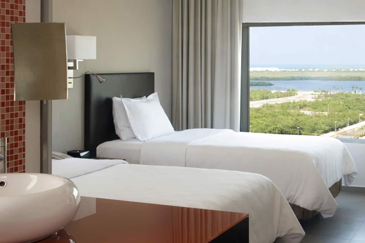 Quarto de hotel com duas camas, uma janela grande com vista para um corpo de água, um banheiro com pia e uma mesa de cabeceira.