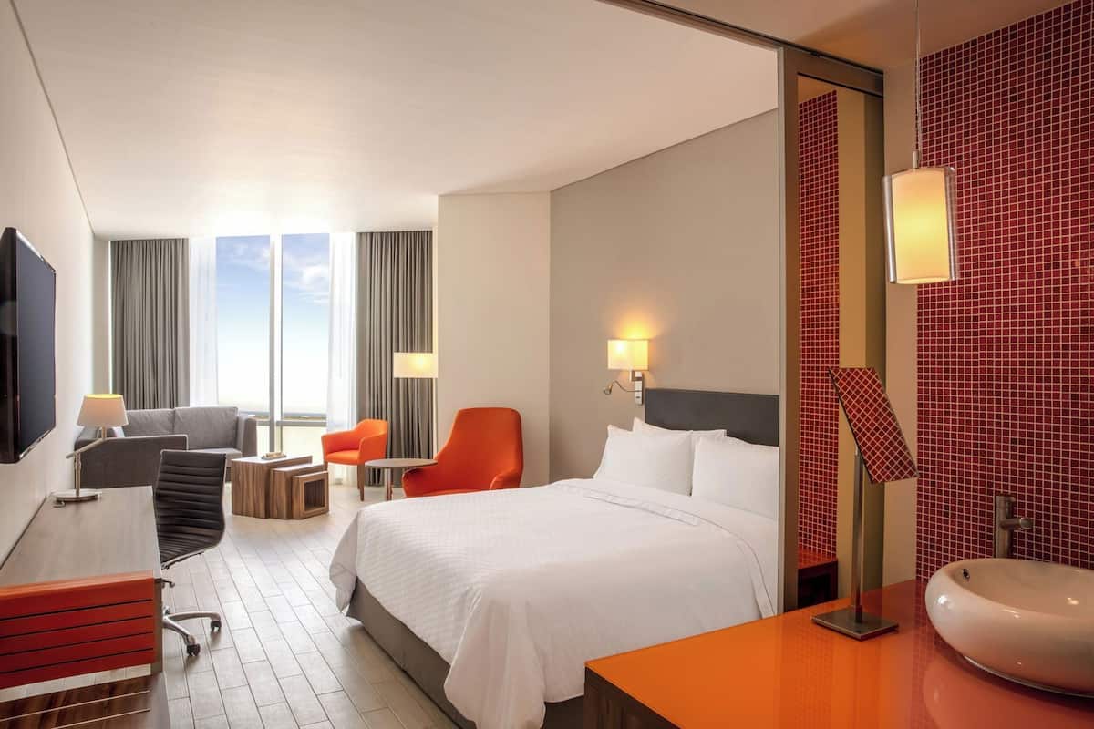 Quarto de hotel com cama, escrivaninha, cadeira, televisão, sofá e banheiro com pia e espelho.