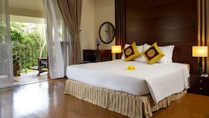 Uma cama grande com roupa de cama branca e duas almofadas decorativas, uma mesa de cabeceira com abajur, um cabeceira de madeira, um espelho redondo e vista para o verde através de uma porta de vidro.