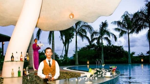 Uma área de bar junto à piscina, com um bartender, uma mulher de vestido roxo e cenário tropical com palmeiras.