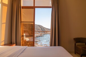 Quarto de hotel com cama, uma cadeira e vista para uma paisagem montanhosa através de uma janela com cortinas.