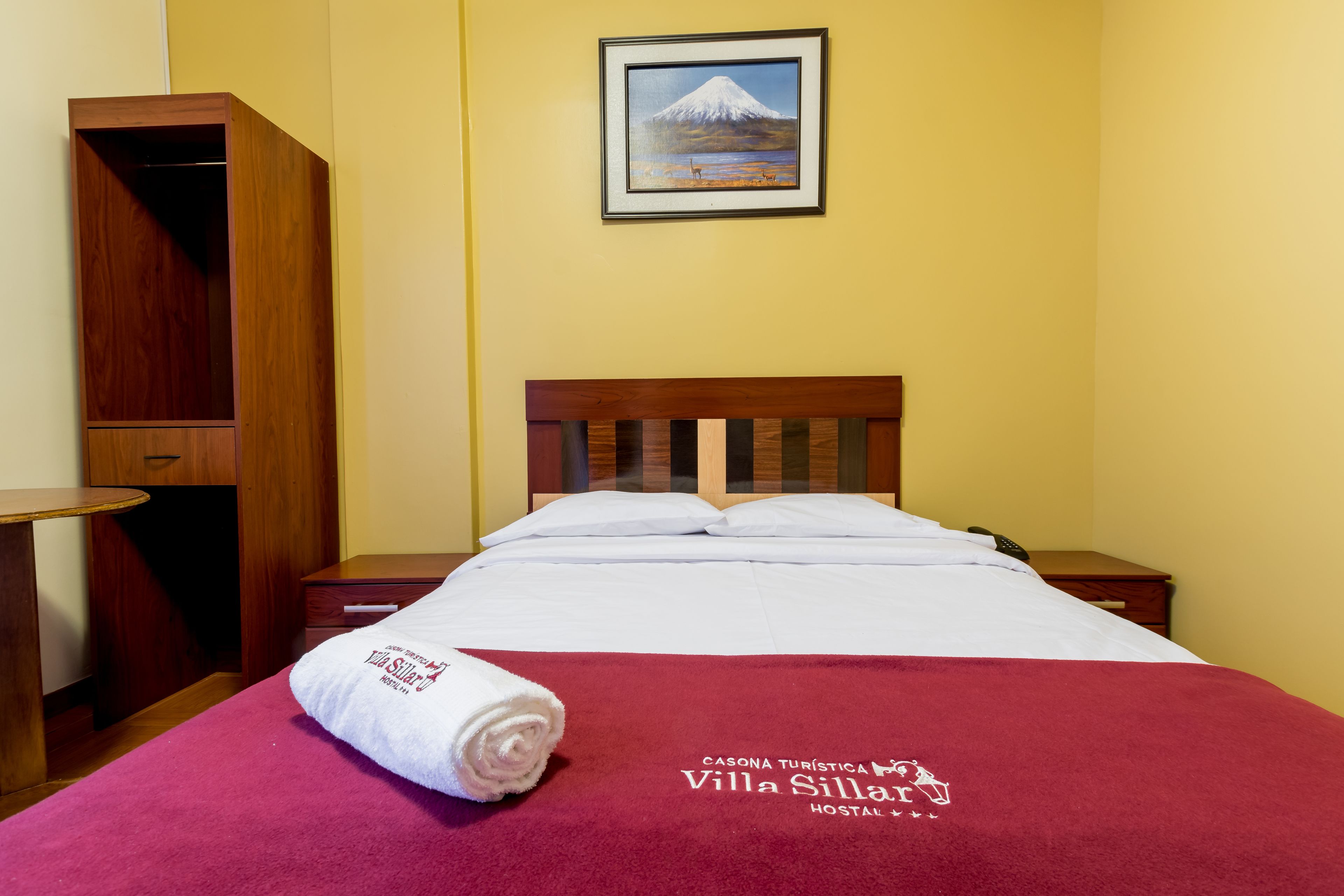 Quarto de hotel com cama, guarda-roupa de madeira, uma pequena mesa e um quadro com uma paisagem de montanha na parede.