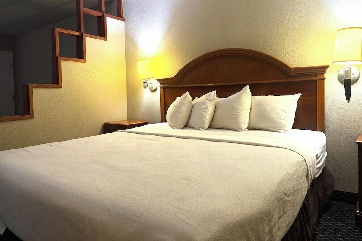 Quarto de hotel com uma cama, cabeceira de madeira, duas luminárias de apoio e uma escada.