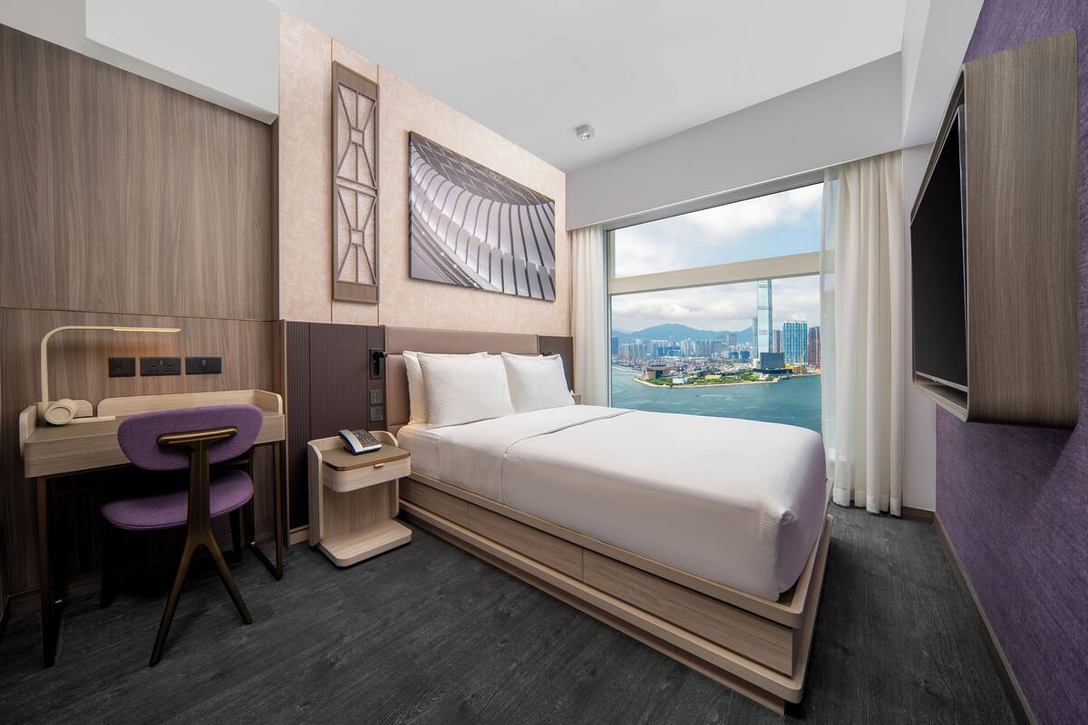 Quarto de hotel moderno com uma cama grande, uma escrivaninha, uma cadeira e vista para a cidade através de uma janela ampla.