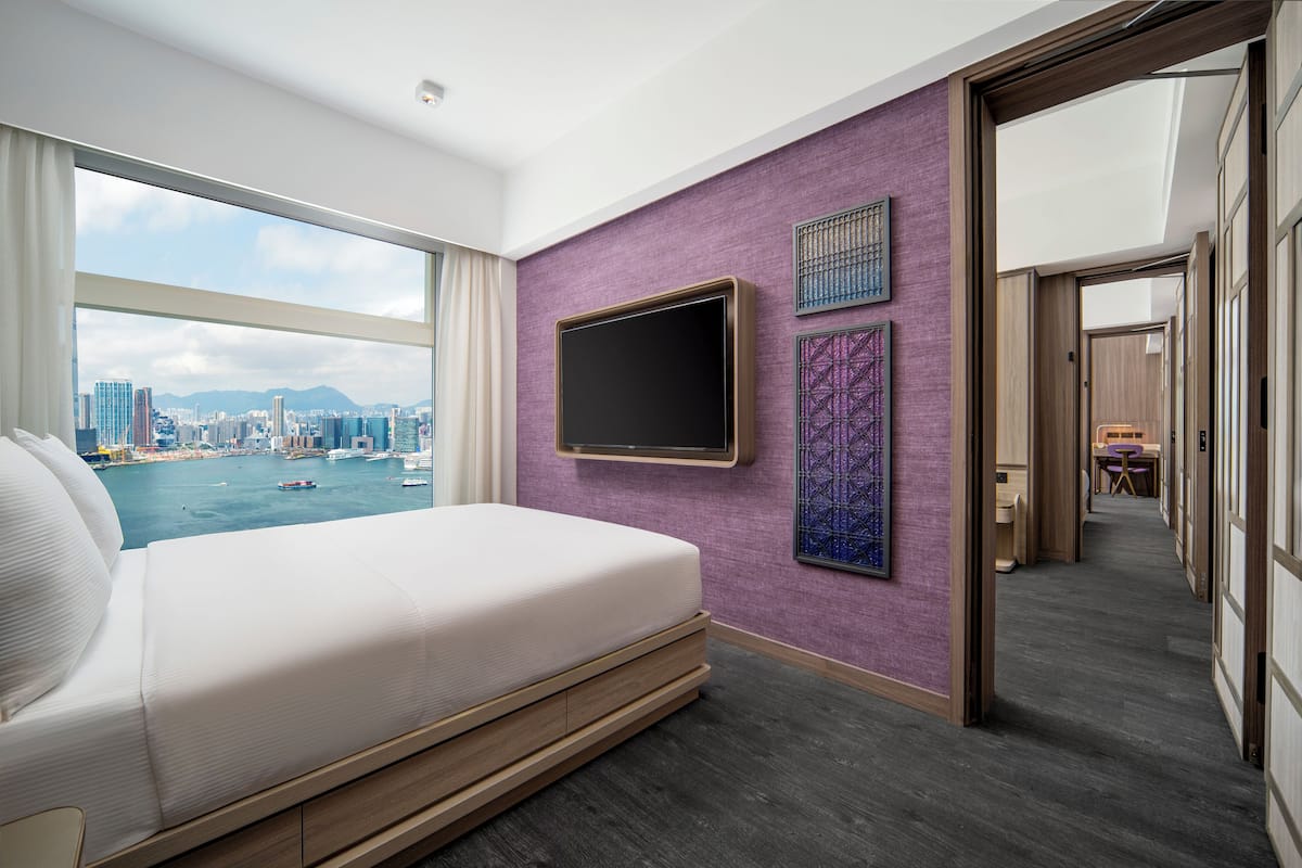 Quarto de hotel moderno com uma cama grande, vista para a cidade, televisão de tela plana e uma pequena mesa com cadeiras.