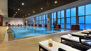 Piscina interna com vista clara do céu através de grandes janelas, área de descanso com assentos e um balcão de recepção.