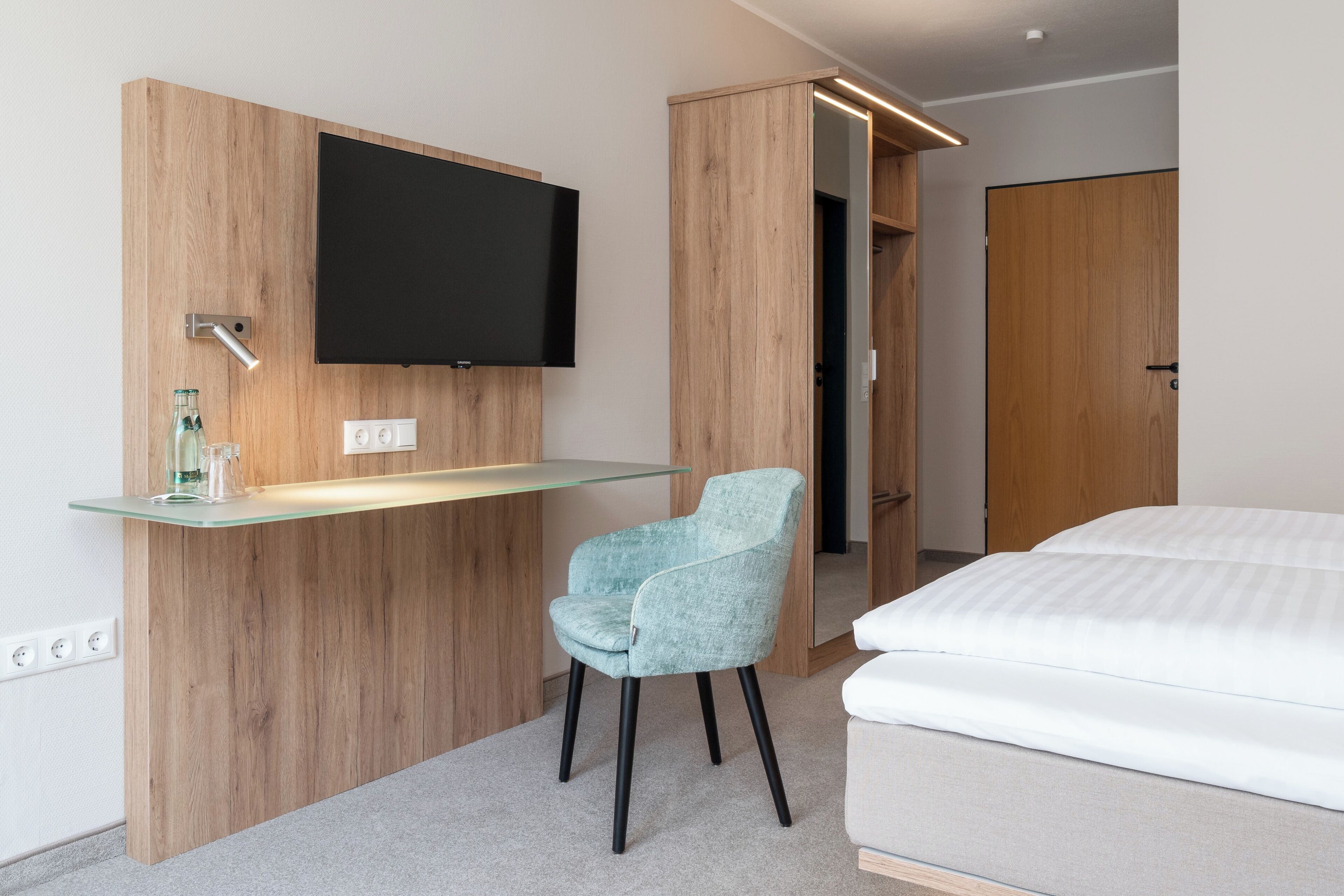 Quarto moderno com guarda-roupa de madeira, mesa de cabeceira de vidro, cadeira azul clara, televisão de tela plana e cama branca.