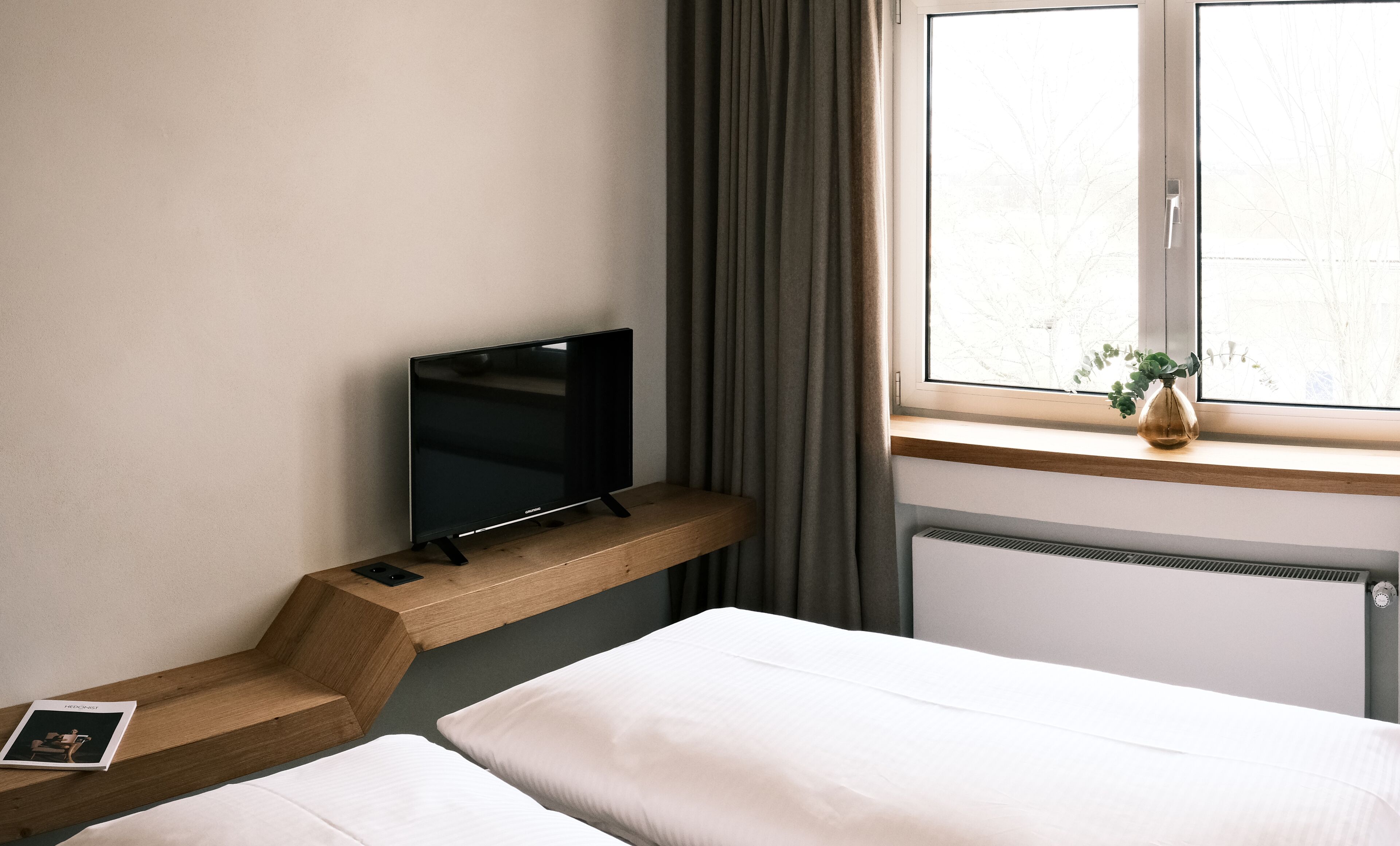 Quarto de hotel com cama, televisão sobre uma estante de madeira, janela com vista para árvores e radiador.