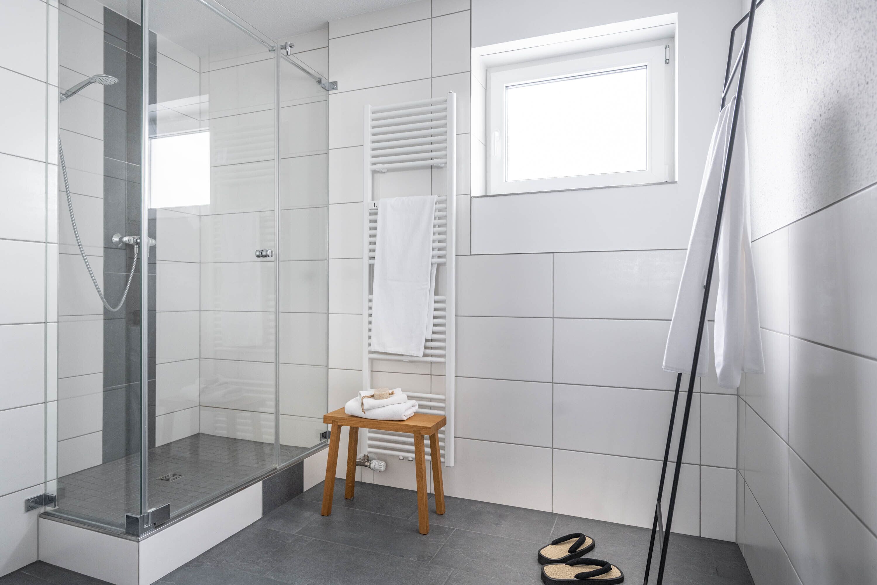 Um banheiro moderno com chuveiro em box de vidro, um banquinho de madeira com toalhas e um radiador branco.