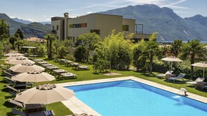 Área de piscina com espreguiçadeiras e guarda-sóis, um edifício moderno e montanhas ao fundo.
