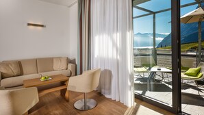Uma sala de estar moderna com sofá, mesa de centro e cadeiras, janelas amplas com cortinas, e vista para montanhas e um lago.