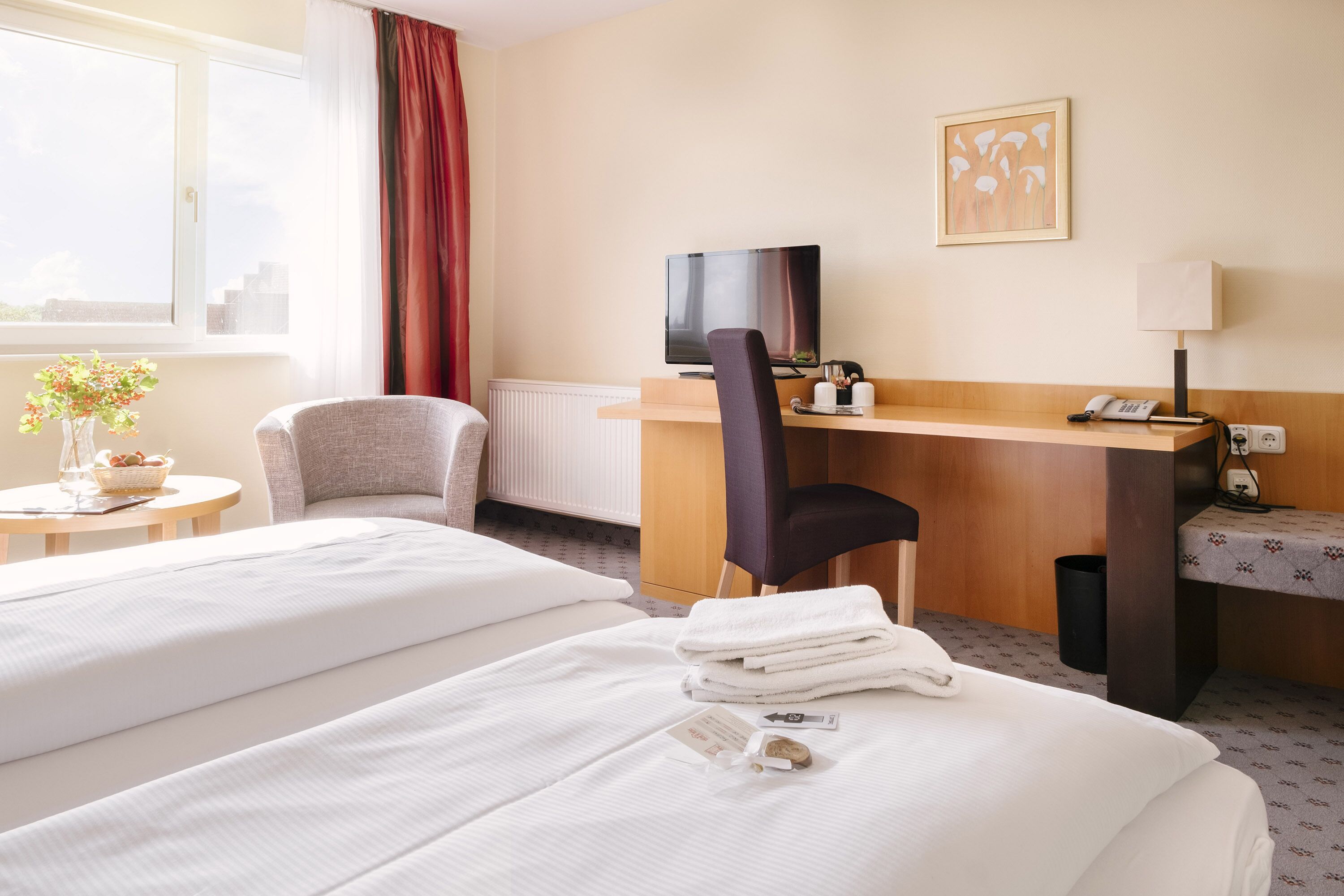 Quarto de hotel com duas camas, uma escrivaninha com cadeira, televisão, abajur e janela com cortinas.